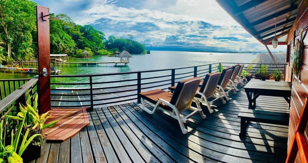 Um deck de madeira com espreguiçadeiras e uma mesa, com vista para um corpo d'água, uma pequena ilha e um bangalô com telhado de palha.