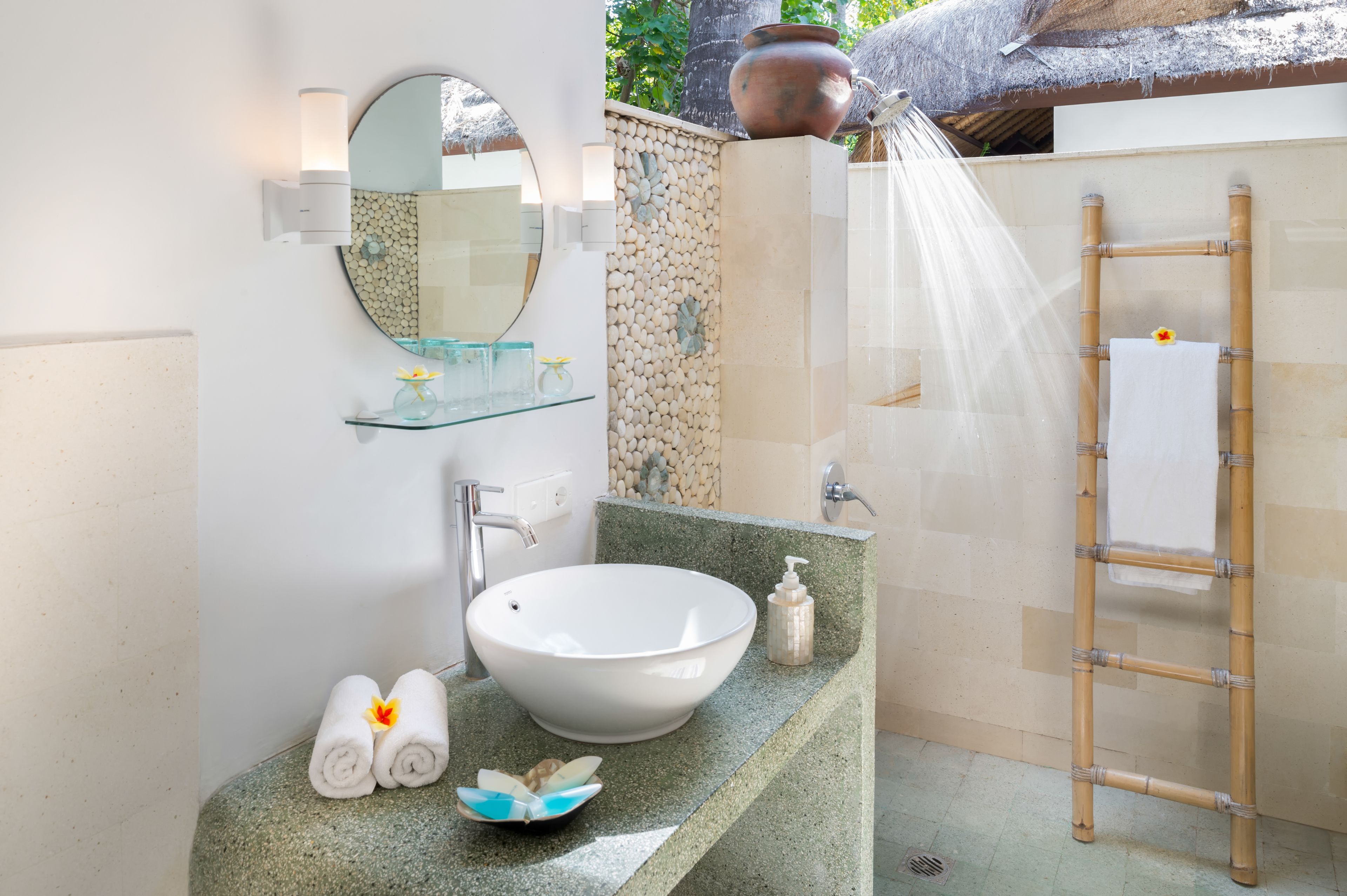 Uma casa de banho com uma grande lavatório branco, um espelho redondo, uma prateleira de vidro com produtos de higiene e um chuveiro com uma escada de bambu.