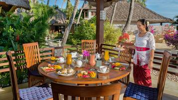 Uma área de refeições ao ar livre tropical com móveis de madeira, uma mesa redonda e uma variedade de frutas e bebidas.