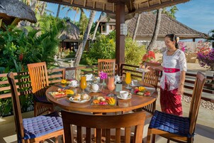 Uma área de refeições ao ar livre tropical com móveis de madeira, uma mesa redonda e uma variedade de frutas e bebidas.