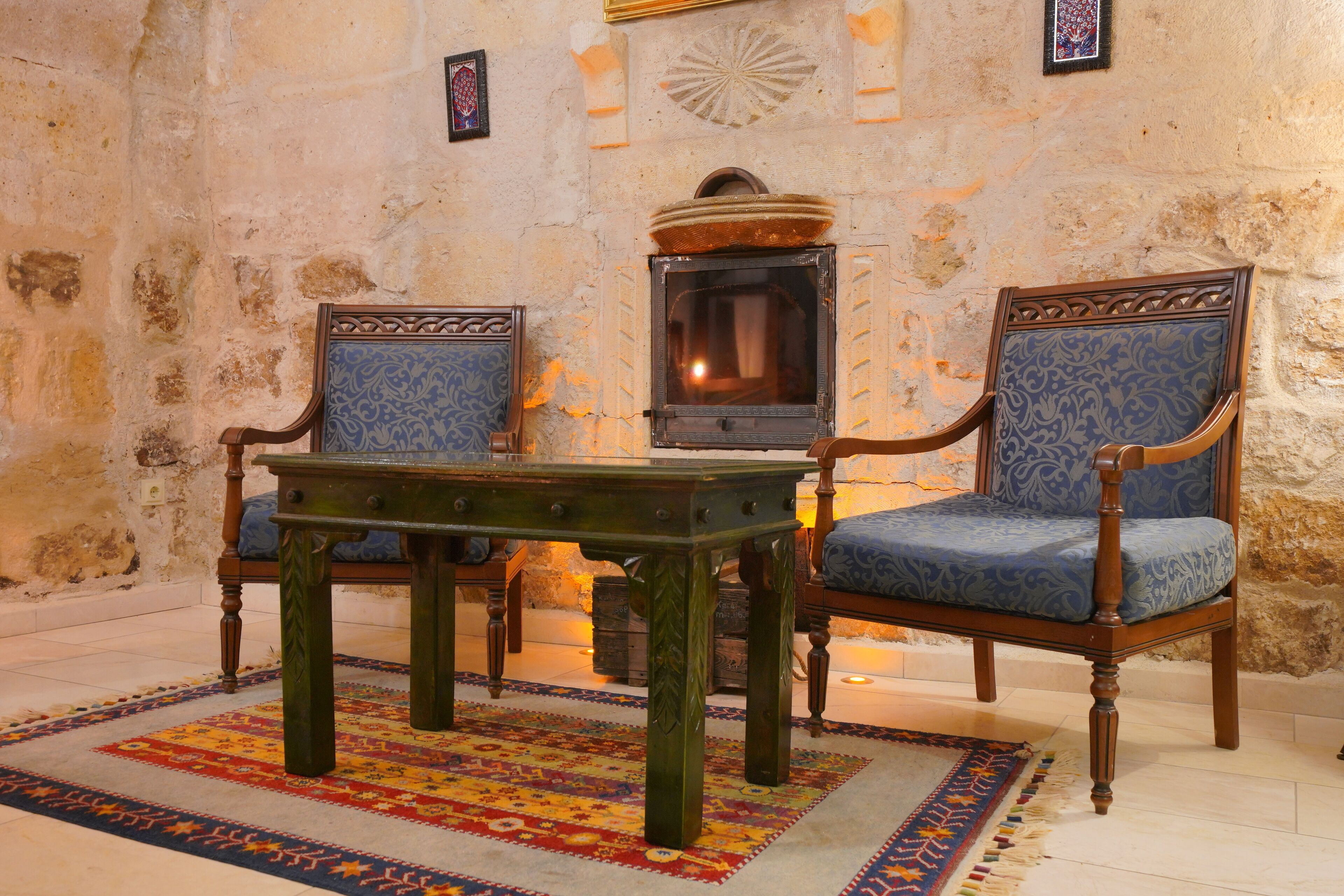 Um quarto com lareira tradicional, duas cadeiras ornamentadas, uma mesa de madeira e um tapete estampado.