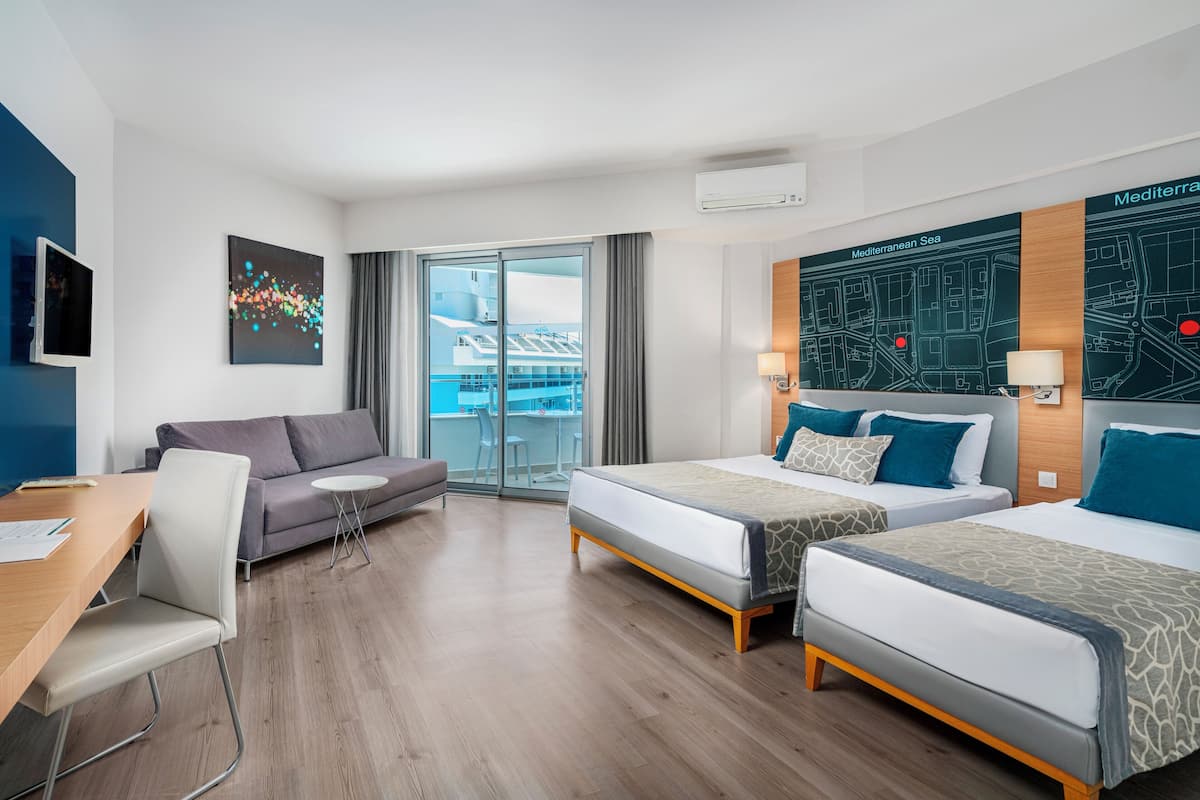 Quarto de hotel com duas camas, um sofá, uma escrivaninha e uma janela grande com vista para o Mar Mediterrâneo.