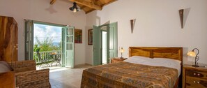 Quarto com cama, cabeceira de madeira, mesa de cabeceira, luminária e vista para o exterior através de uma porta aberta.