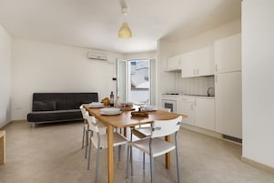 Uma cozinha compacta com armários brancos, uma mesa de jantar com cadeiras e um sofá ao fundo.