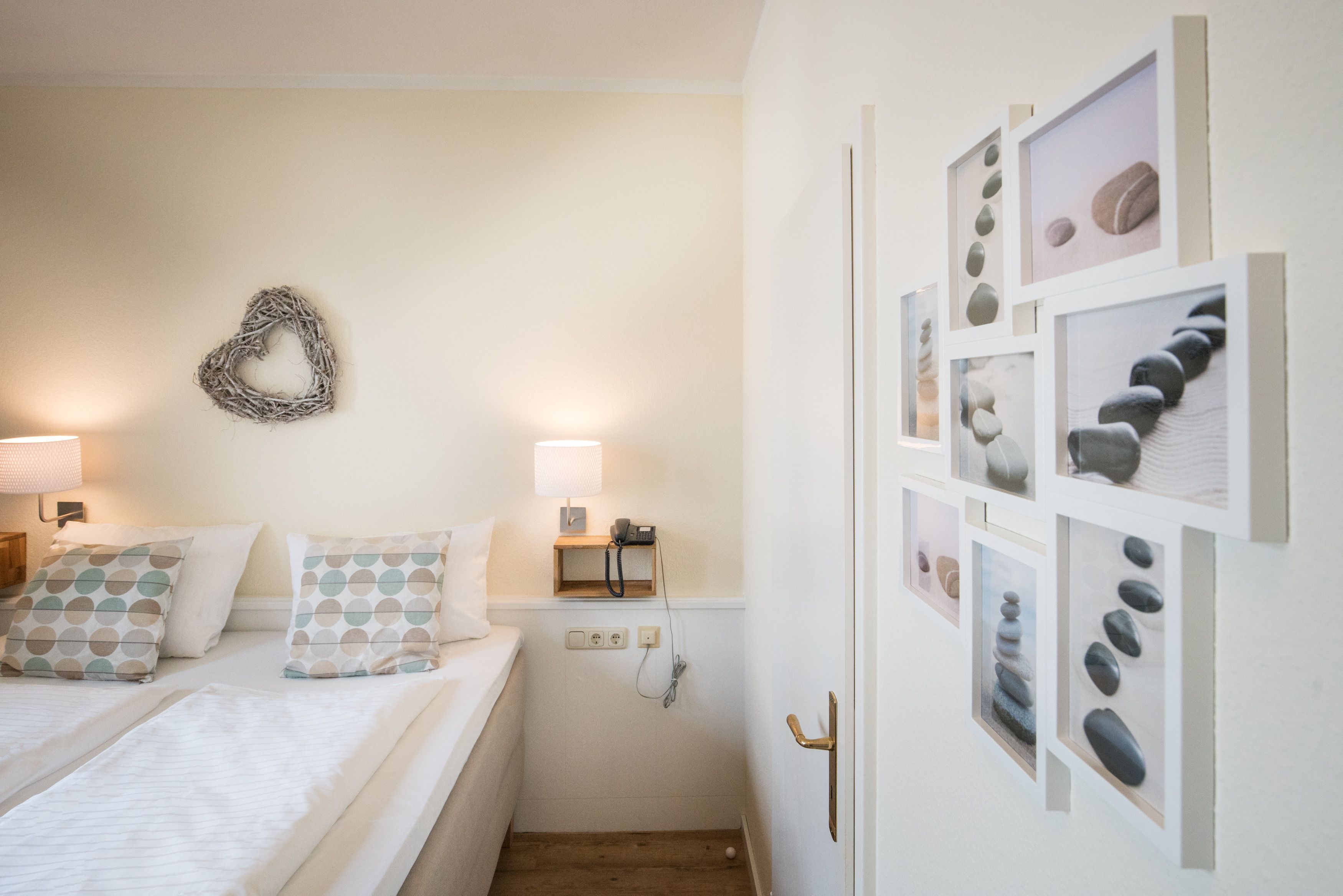 Quarto com cama, almofadas, luminária na parede, prateleira pequena com um telefone e quadros na parede.
