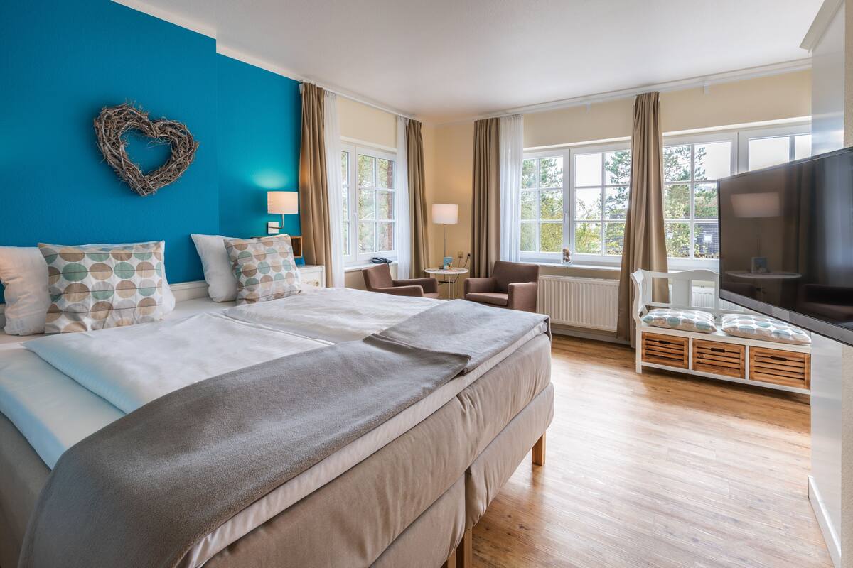 Quarto de hotel com uma cama grande, televisão, escrivaninha com cadeira e vista para o exterior pelas janelas.