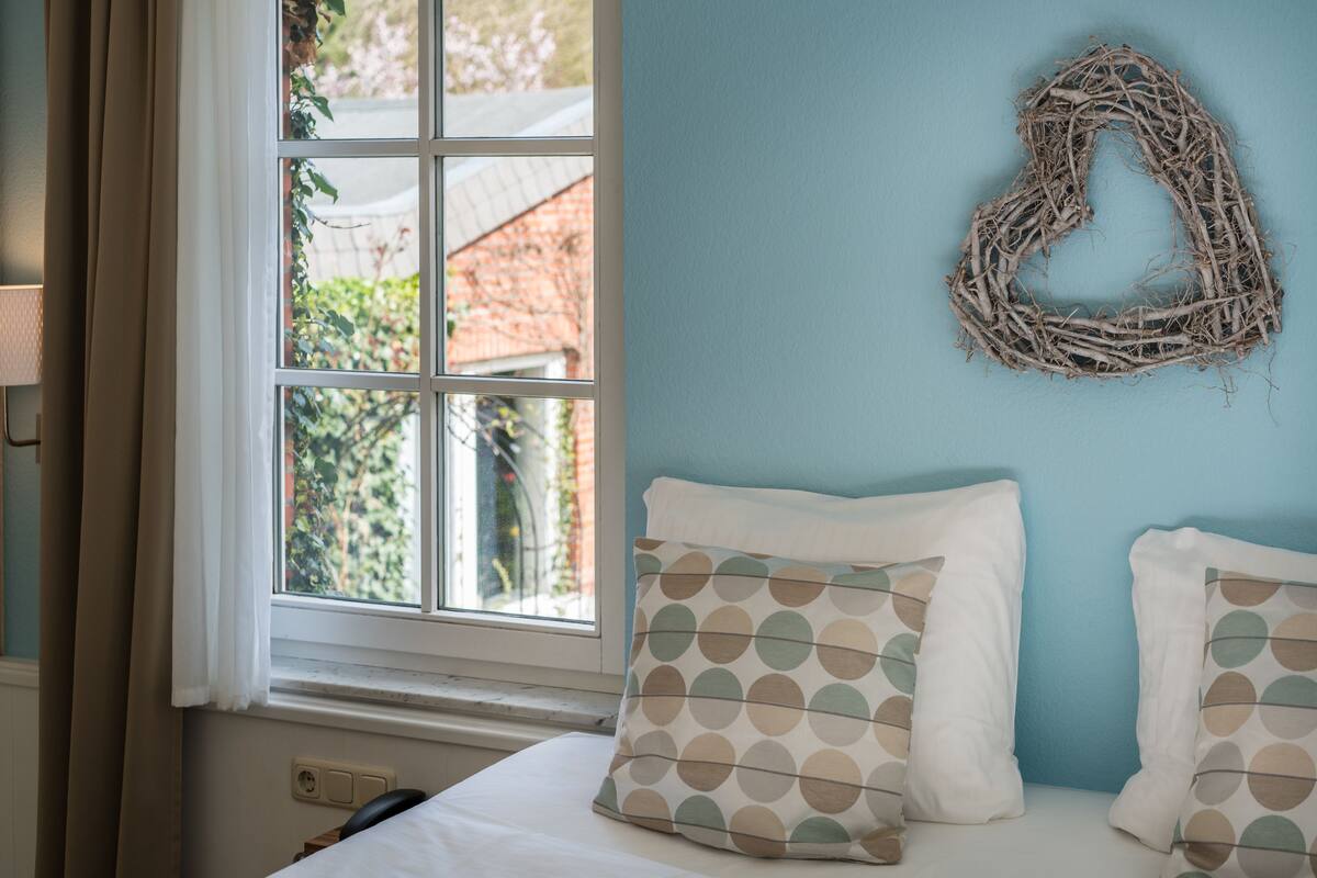 Quarto com cama, duas almofadas estampadas com poás, decoração de parede em tear e janela com vista para vegetação e um edifício.
