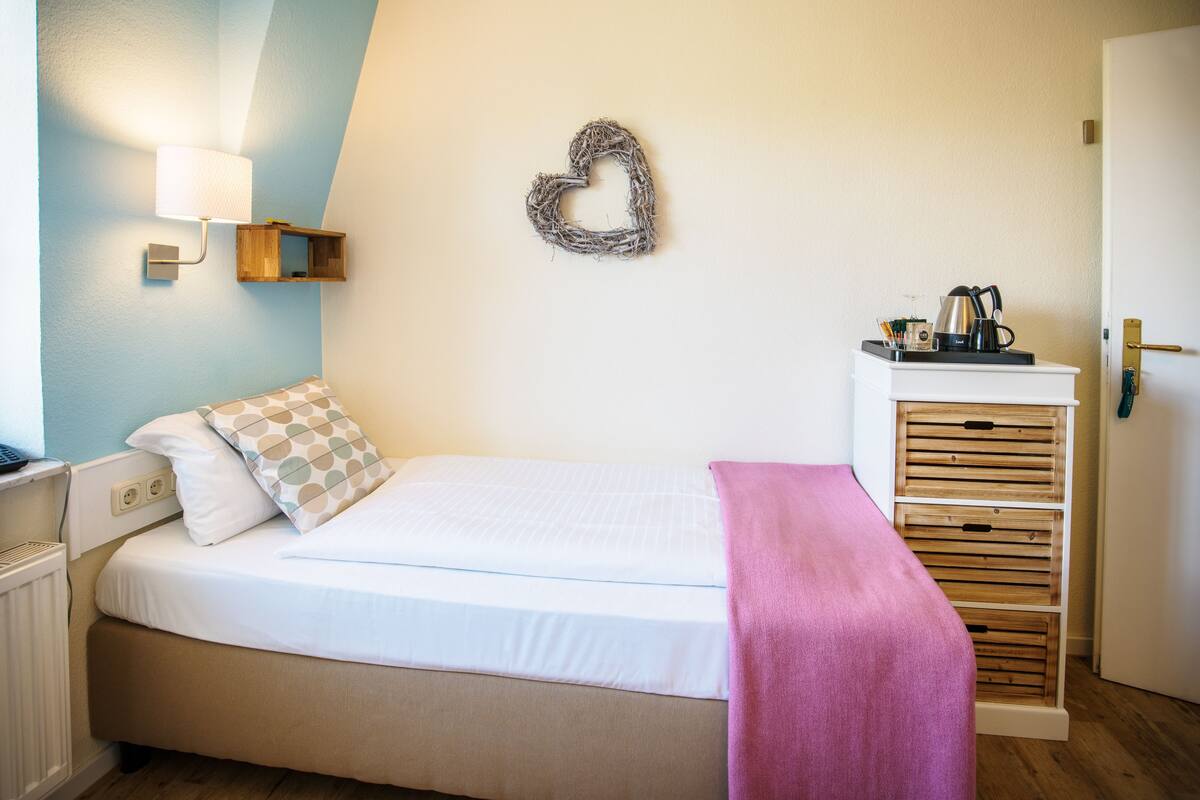 Cama bem arrumada com travesseiro quadriculado em branco e bege, manta rosa e decoração em formato de coração na parede.