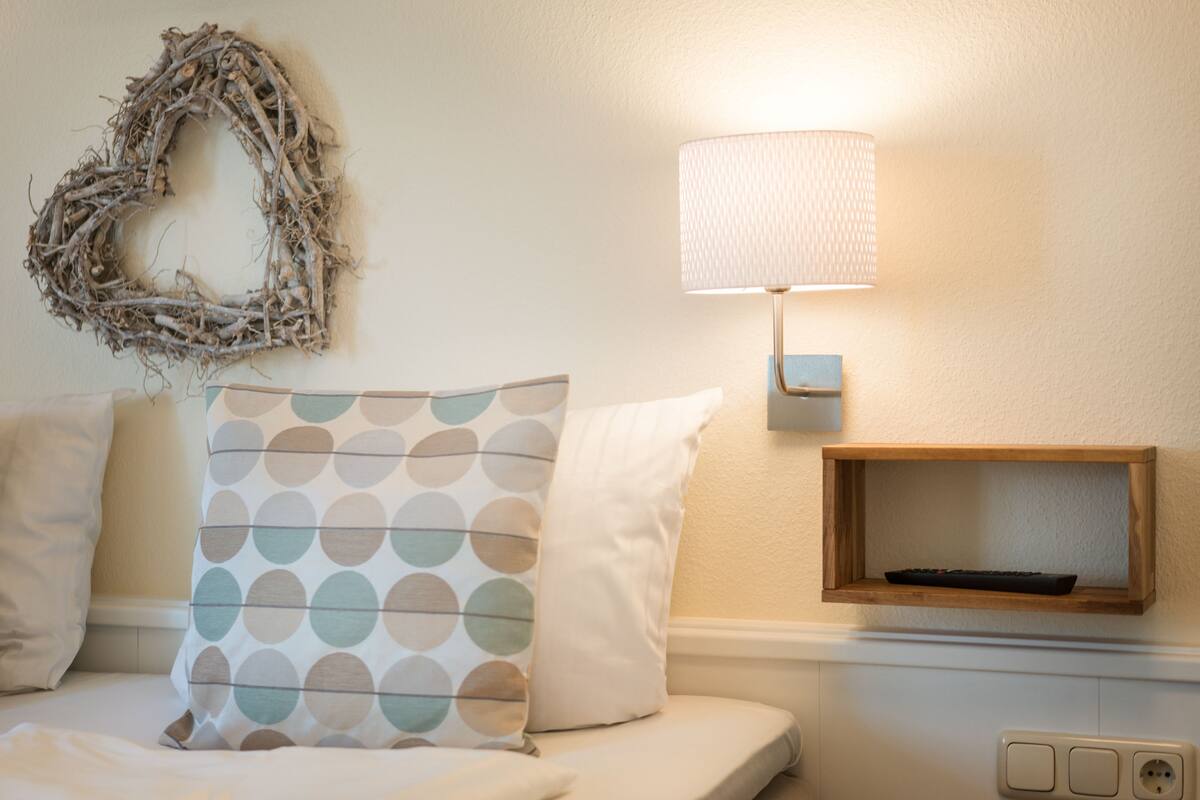 Cama bem arrumada com almofadas estampadas, luminária na parede e prateleira com controle remoto.