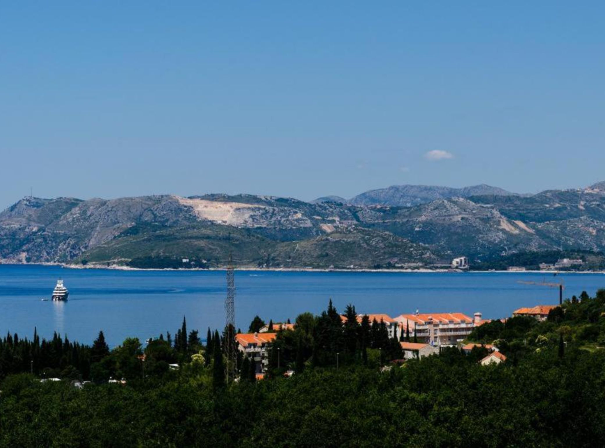 Vista costeira com mar azul límpido, um iate e montanhas ao fundo.