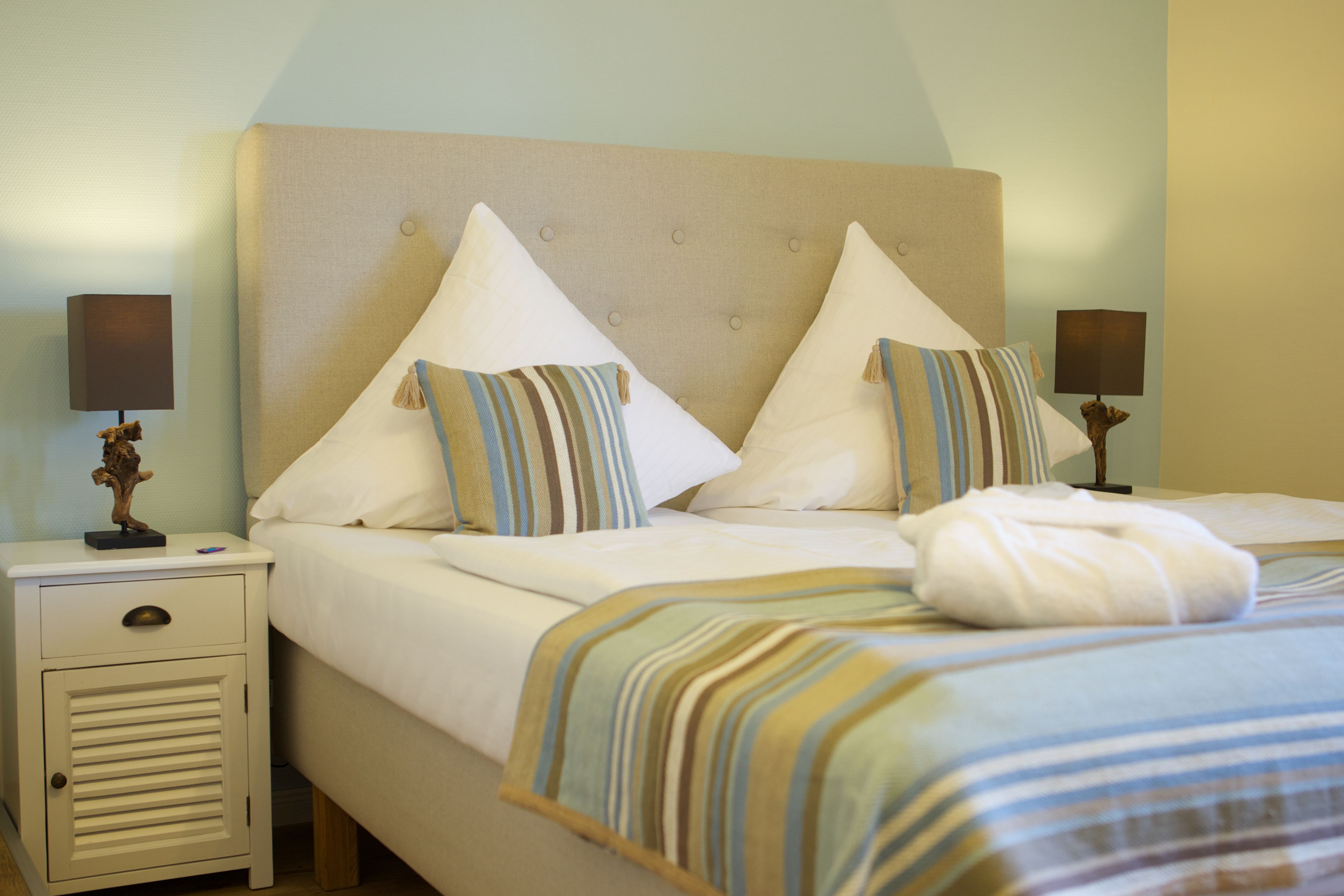 Cama bem arrumada com roupa de cama listrada, duas luminárias de cabeceira e uma mesa de cabeceira com gaveta.