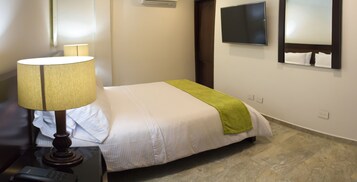 Uma cama bem arrumada com um cobertor verde, uma luminária de cabeceira, uma televisão de tela plana e um espelho na parede.