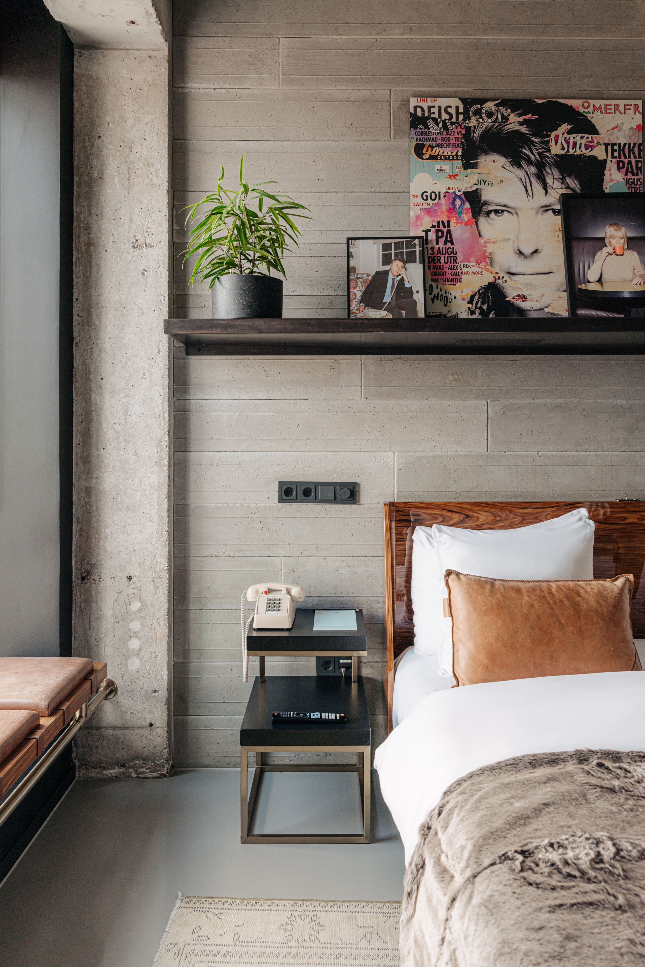 Quarto com cama, mesa de cabeceira com telefone, pequena mesa com abajur, prateleira na parede com planta e cartazes, e parede de concreto.
