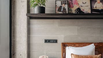 Quarto com cama, mesa de cabeceira com telefone, pequena mesa com abajur, prateleira na parede com planta e cartazes, e parede de concreto.