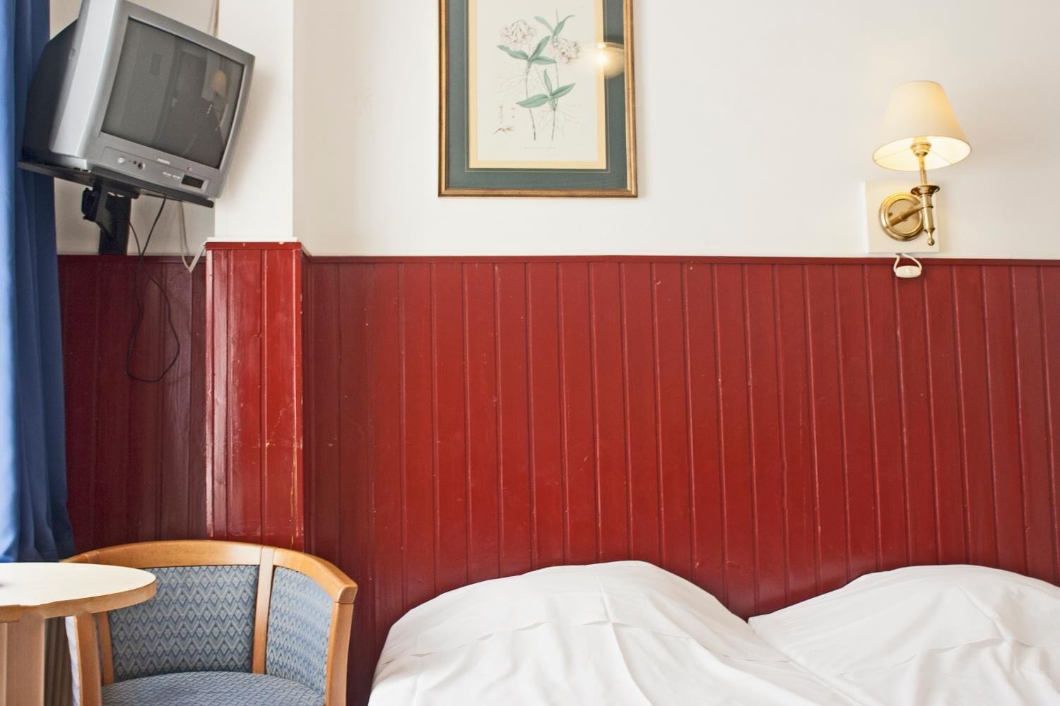 Quarto de hotel com painel vermelho, uma cama de solteiro, uma mesinha, uma cadeira, uma televisão e uma luminária fixada na parede.