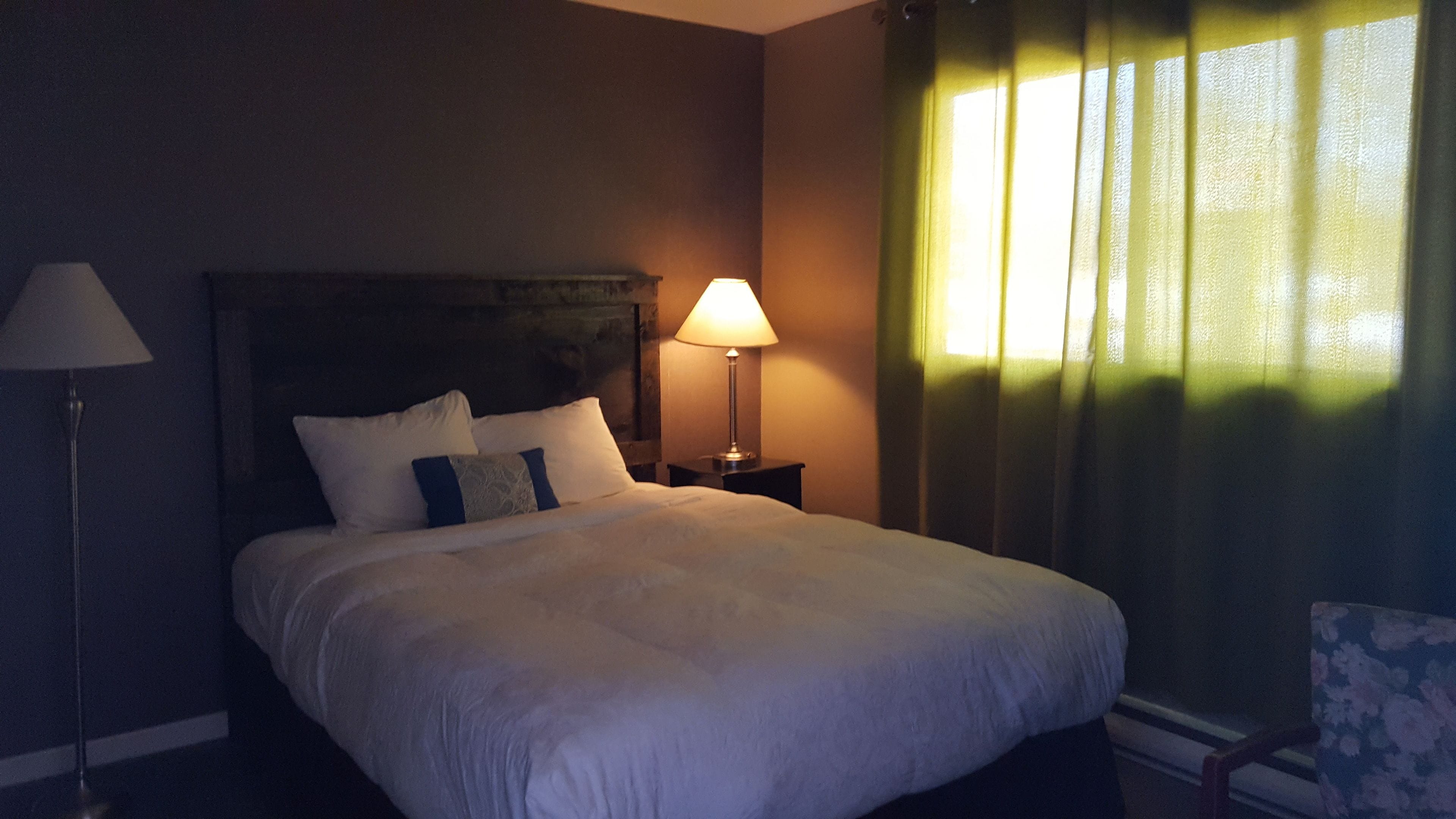 Uma cama bem arrumada com roupa de cama branca, cabeceira e duas luminárias de cabeceira.