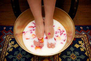 Uma bacia com os pés de uma pessoa submersos em um líquido leitoso, com pétalas de rosa, sobre um tapete estampado.