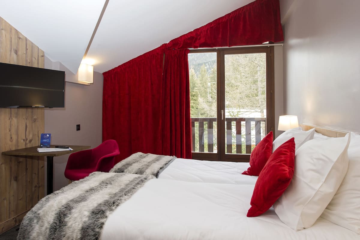 Quarto de hotel com cama, cortinas vermelhas, televisão, escrivaninha e uma cadeira.