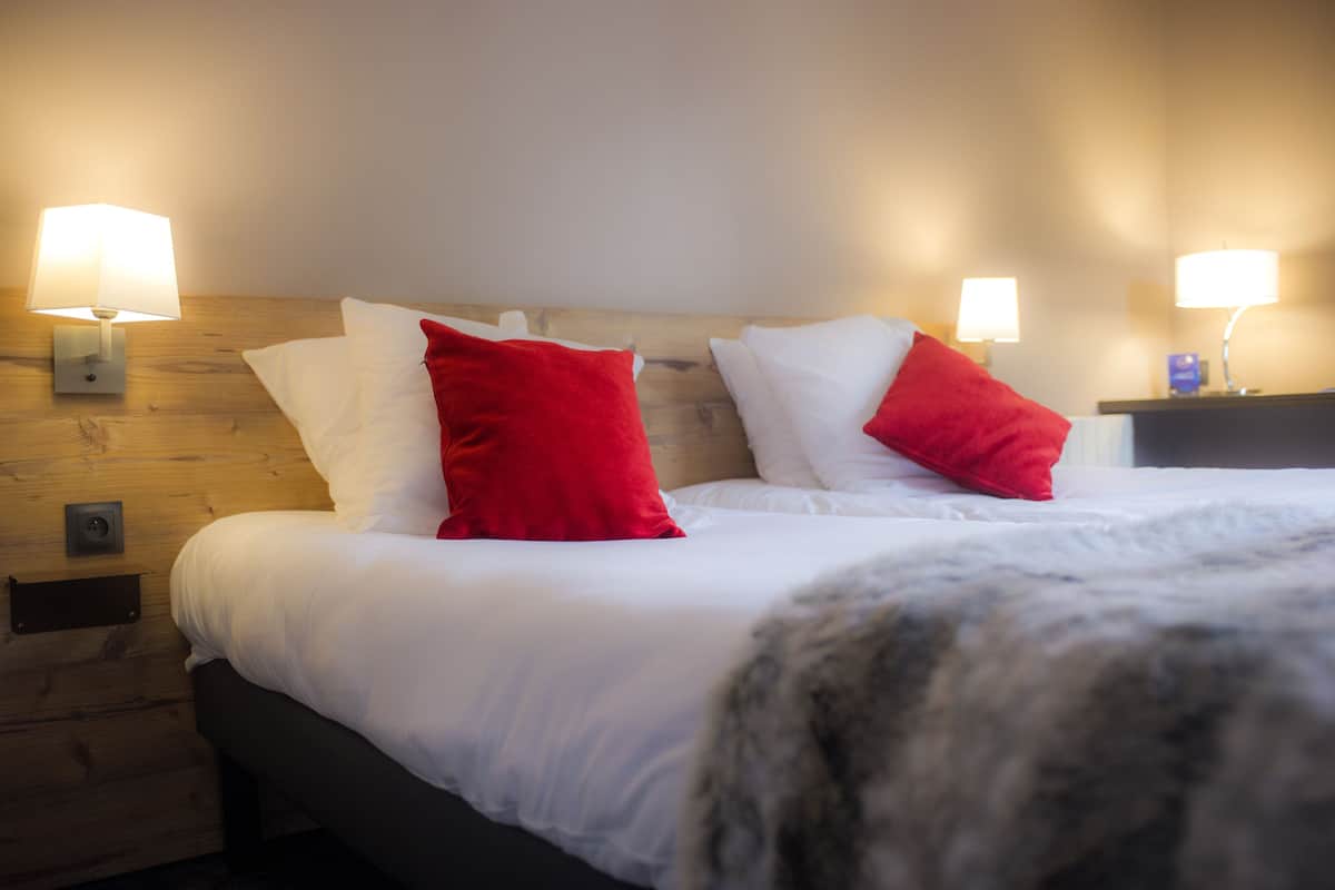 Cama bem arrumada com almofadas brancas e vermelhas, duas luminárias de cabeceira e uma cabeceira de madeira.