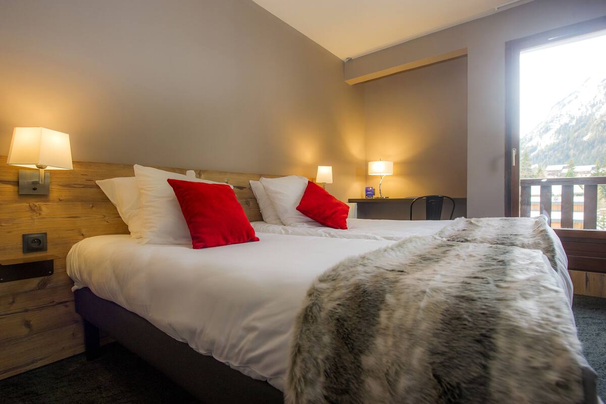 Quarto de hotel com uma cama grande, duas luminárias de cabeceira, uma cabeceira de madeira e vista para as montanhas pela janela.
