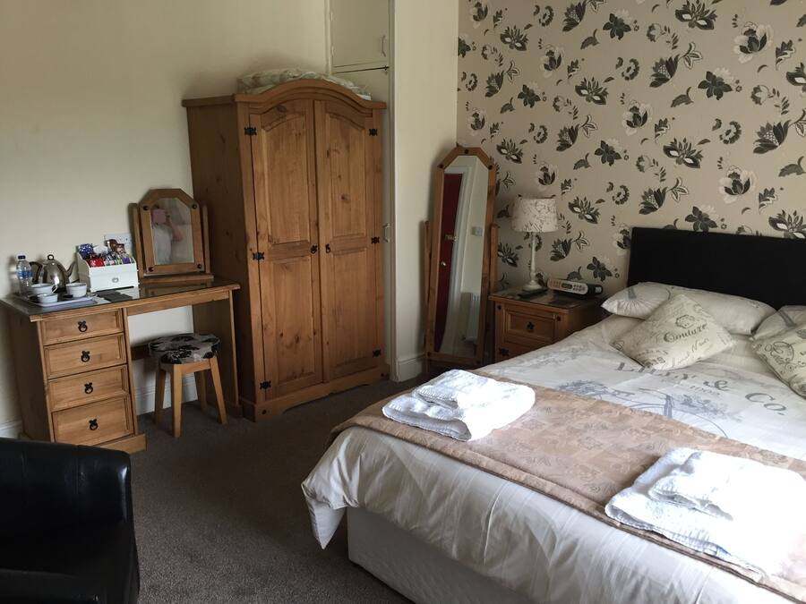 Quarto com cama, guarda-roupa de madeira, escrivaninha com gavetas, cadeira, espelho e mesa de cabeceira com abajur.