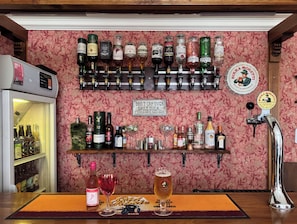Uma área de bar com prateleiras cheias de garrafas de bebidas, uma torneira de chopp e uma placa com a inscrição 'NÃO SE ESTRESSE POR LEITE DERRAMADO. PODIA TER SIDO CERVEJA'.