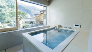 Um banheiro moderno com uma banheira grande, uma janela com vista para um edifício tradicional e montanhas, e um aquecedor de água na parede.