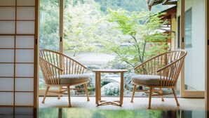 Um quarto japonês tradicional com tatames, duas cadeiras de madeira, uma mesinha e vista para um jardim através de um biombos shoji.