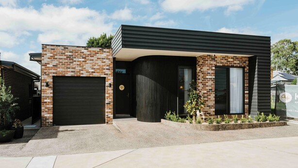 Uma casa moderna de dois andares com fachada de tijolos, porta de garagem escura e um jardim frontal bem cuidado.