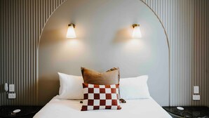 Quarto de hotel com uma cama, duas luminárias de cabeceira, um travesseiro xadrez e um aparelho de luz fixado na parede.