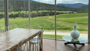 Uma área de refeições com uma mesa grande e cadeiras, uma mesa de vidro, e vista para um vinhedo e montanhas através de grandes janelas.