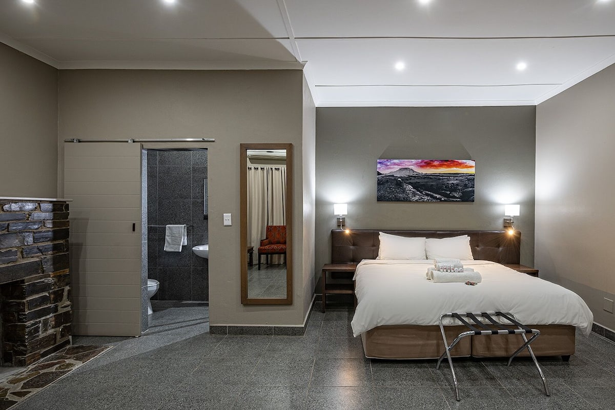Quarto de hotel com lareira de pedra, cama com roupa de cama branca, mesa de cabeceira com luminária e um quadro na parede.