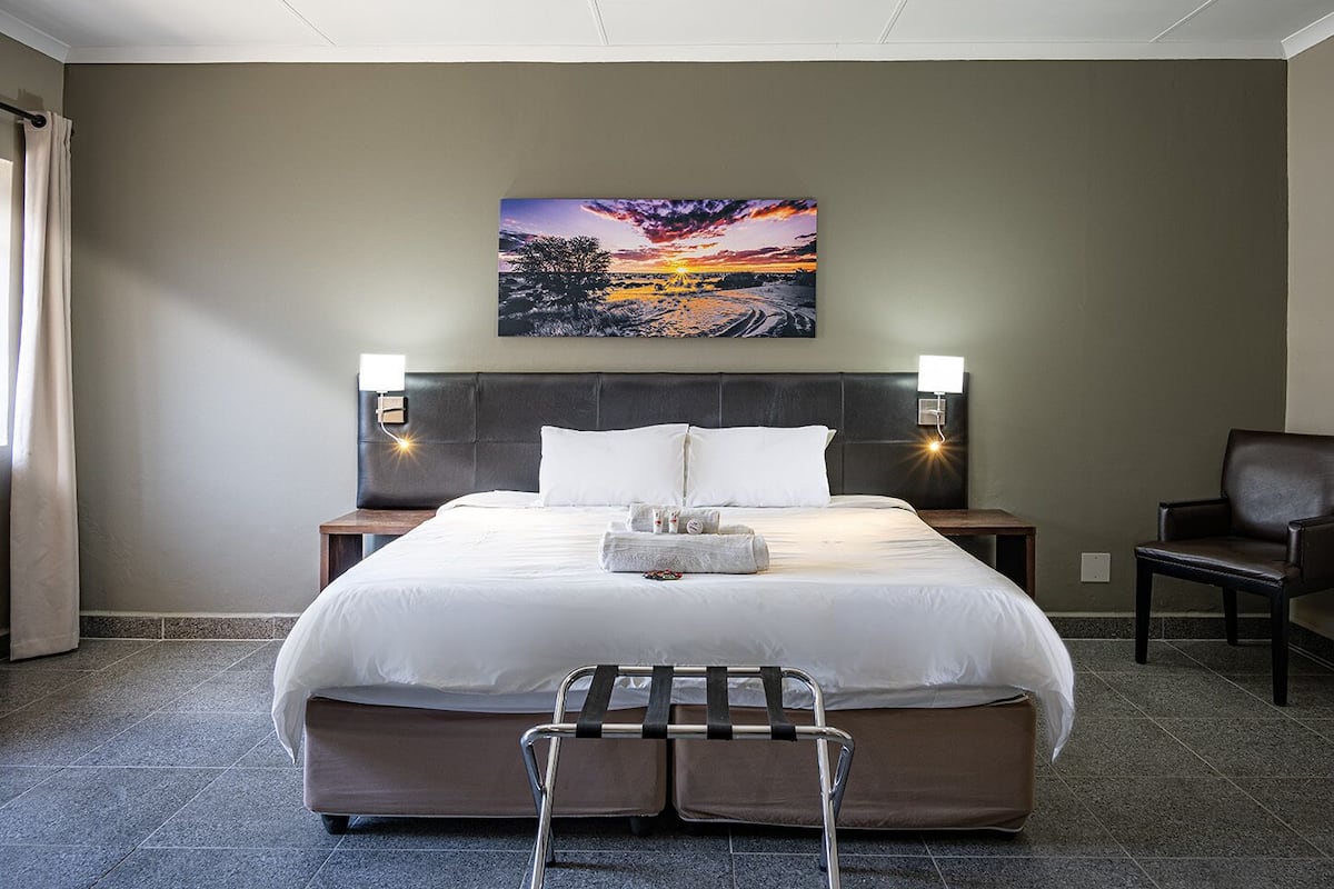Quarto de hotel com uma cama grande, duas mesas de cabeceira, uma cadeira, um quadro na parede e uma janela com cortinas.