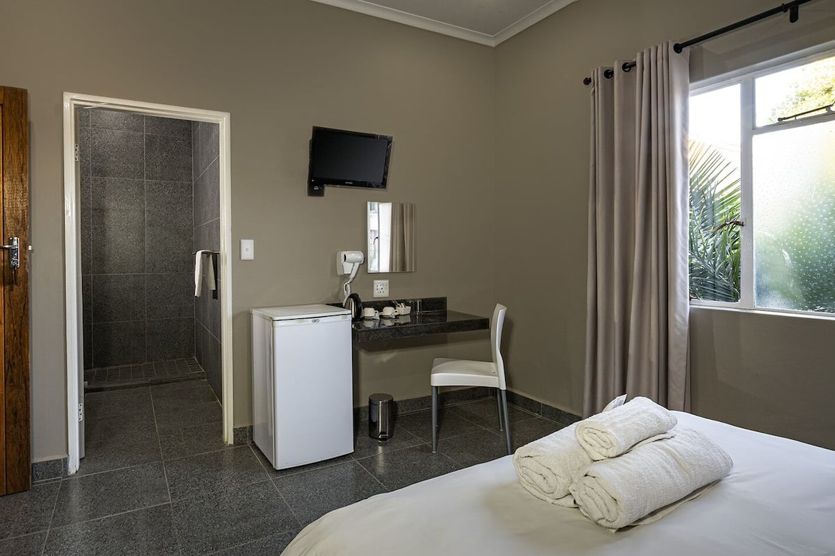 Quarto de hotel com cama, escrivaninha com cadeira, pequeno frigorífico, área de chuveiro e televisão na parede.