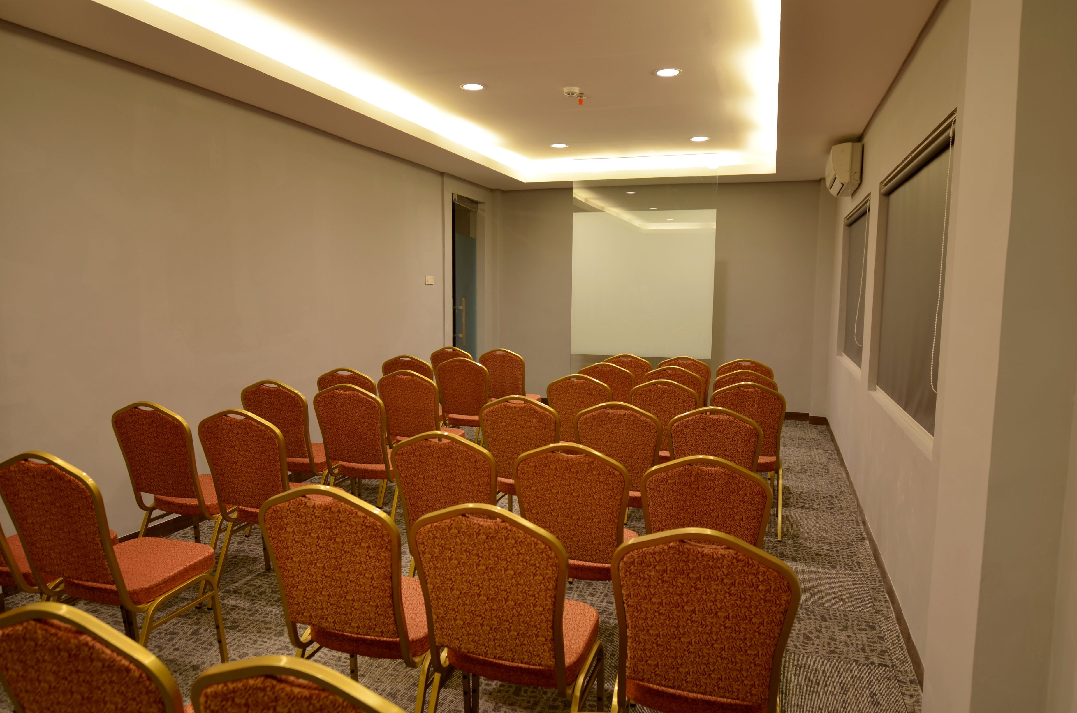 Uma sala de conferências com fileiras de cadeiras laranja e uma tela grande na frente.