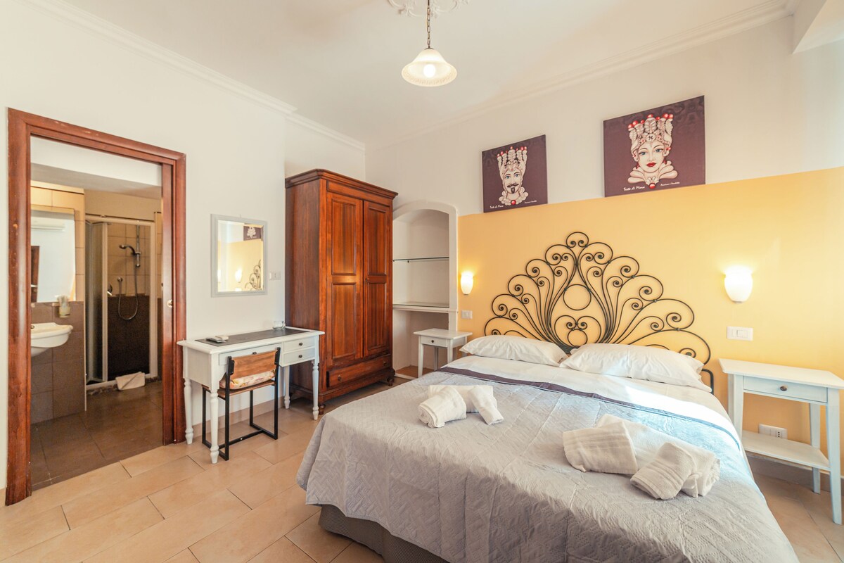 Quarto com cama, escrivaninha, cadeira, guarda-roupa e banheiro visível por uma porta aberta.