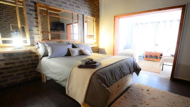 Quarto com cama grande, cabeceira de madeira, mesinha com abajur e vista para o exterior por uma porta deslizante.