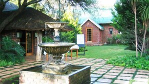 Um jardim com uma fonte de pedra, uma construção com telhado de palha e um edifício de tijolo com telhado verde.