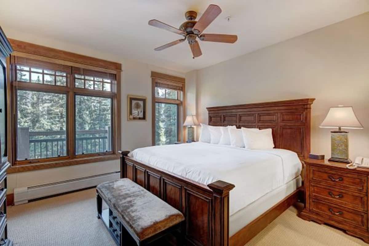 Quarto com uma cama grande, cabeceira de madeira, duas mesas de cabeceira com luminárias, um banquinho e um ventilador de teto.