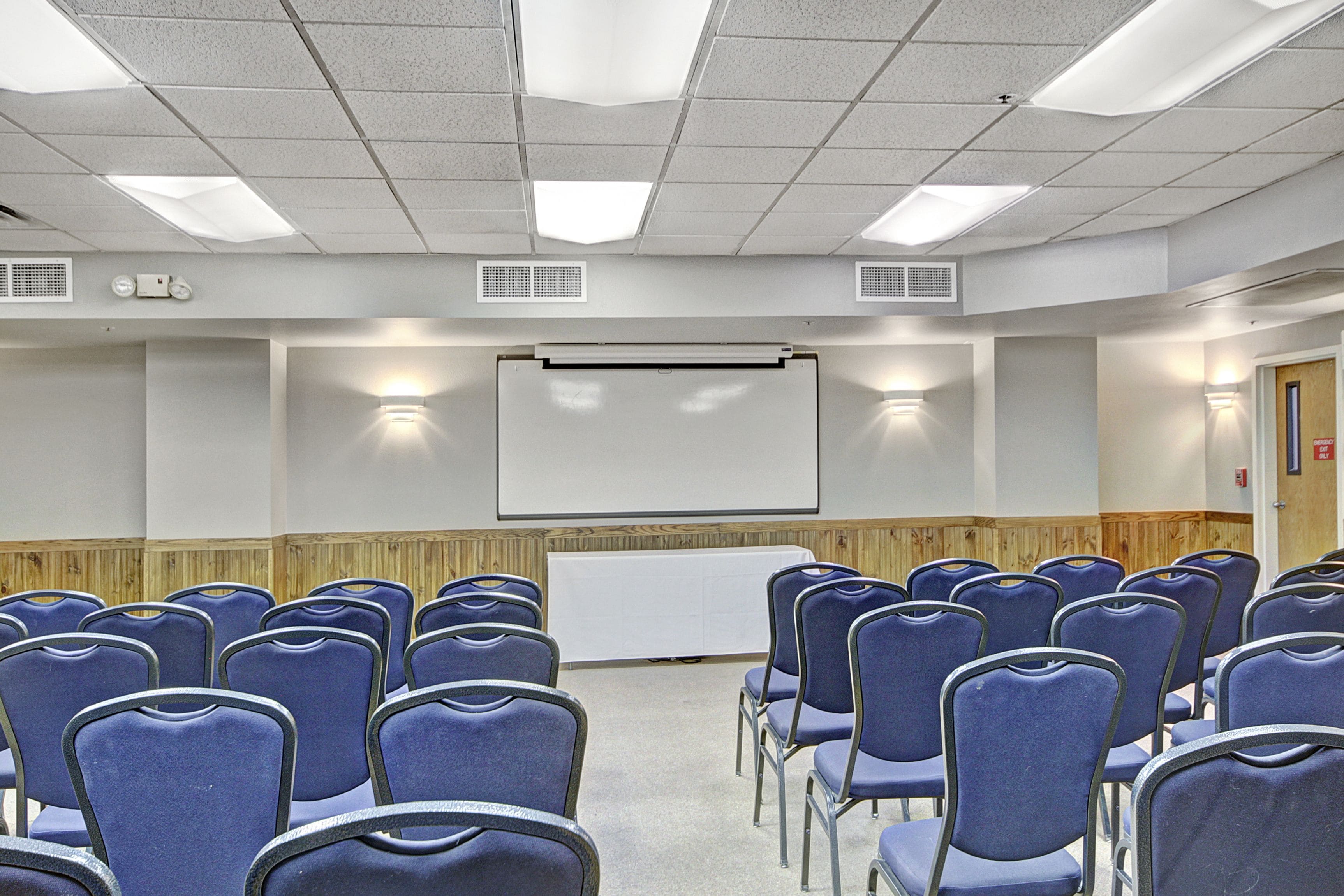Uma sala de conferência com fileiras de cadeiras azuis, uma tela de projeção branca e iluminação embutida.