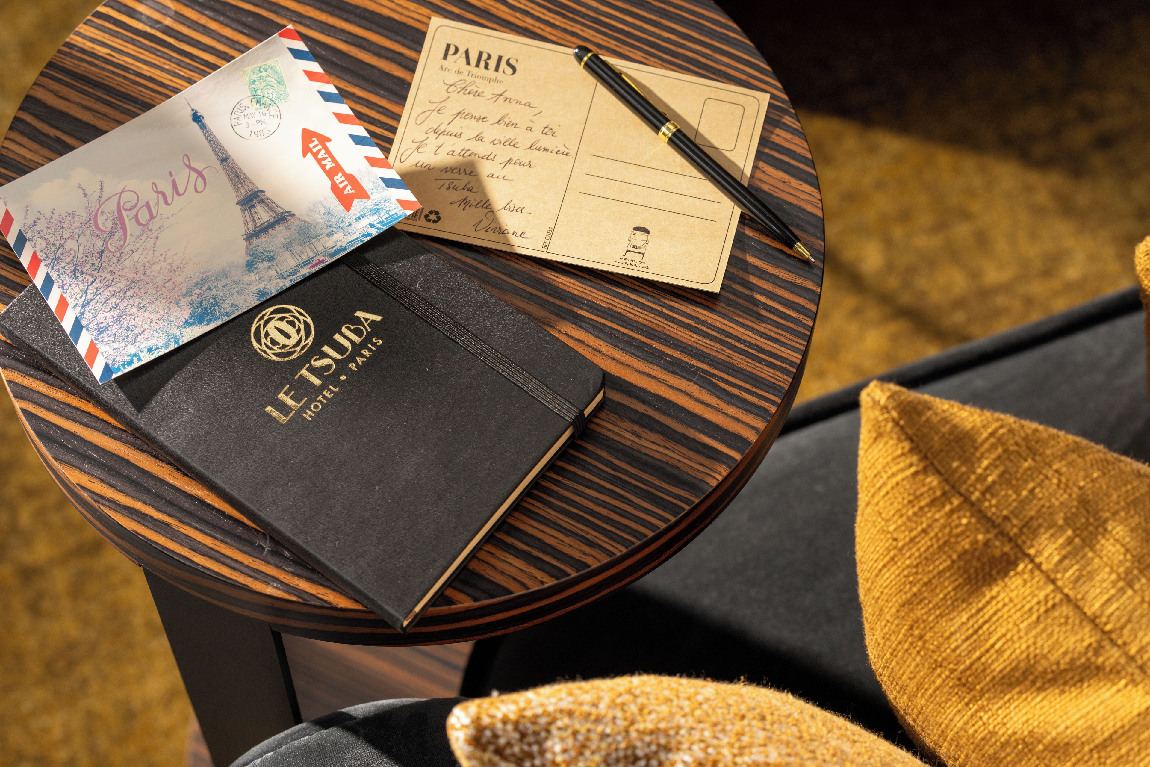 Uma mesa redonda de madeira com um caderno preto, um postal, uma caneta e um pequeno envelope.