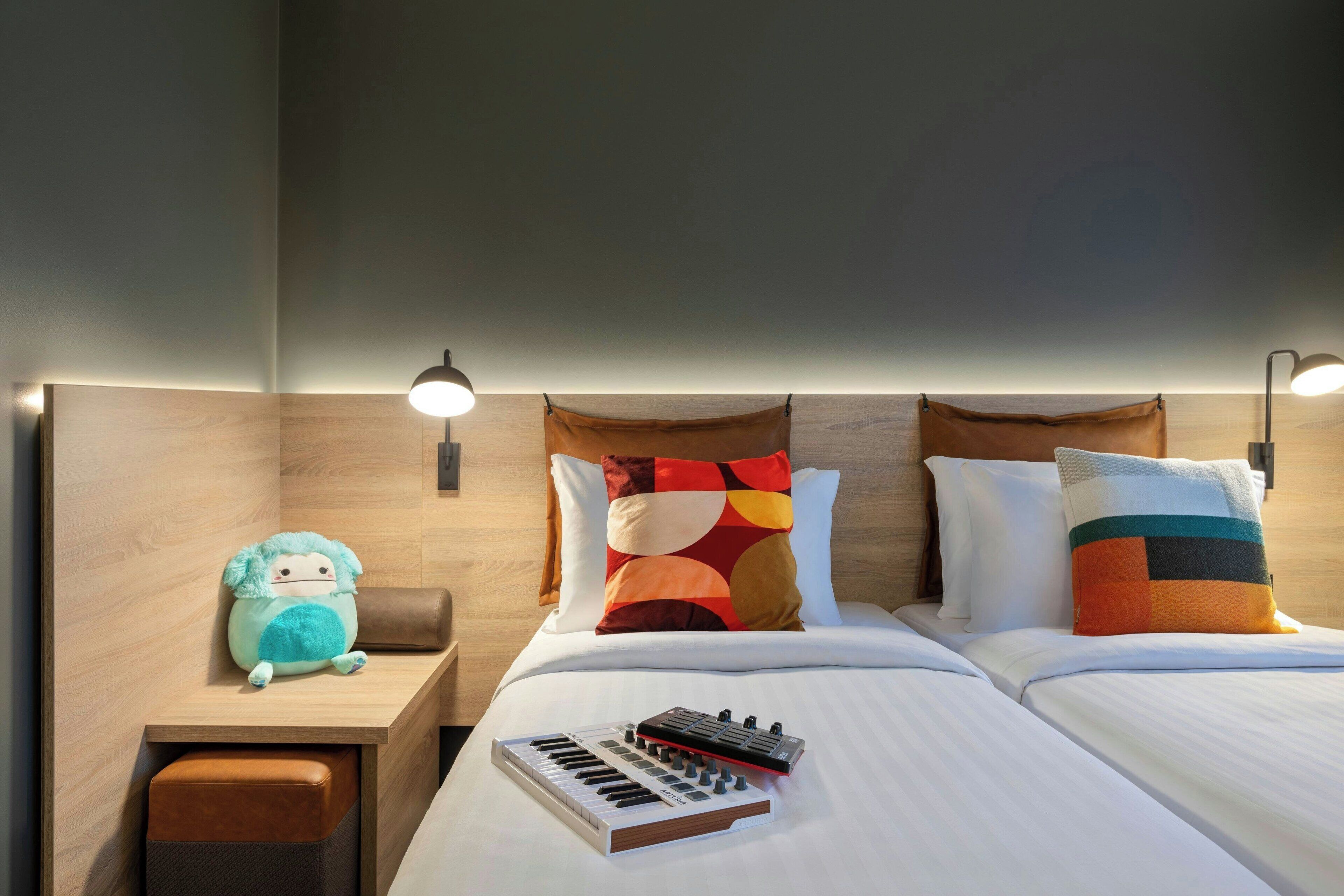 Quarto de hotel com duas camas, uma escrivaninha, um brinquedo de pelúcia e um teclado.