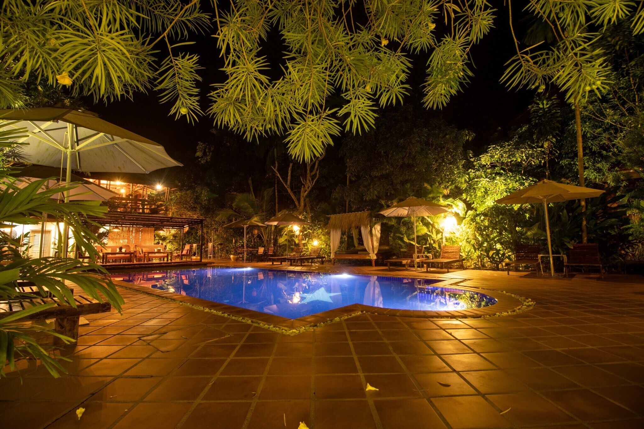 Uma área de piscina à noite, com chão ladrilhado, guarda-sóis e mobiliário de exterior.