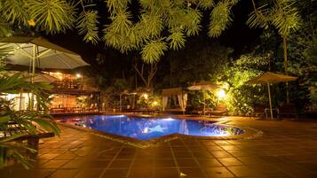Uma área de piscina à noite, com chão ladrilhado, guarda-sóis e mobiliário de exterior.