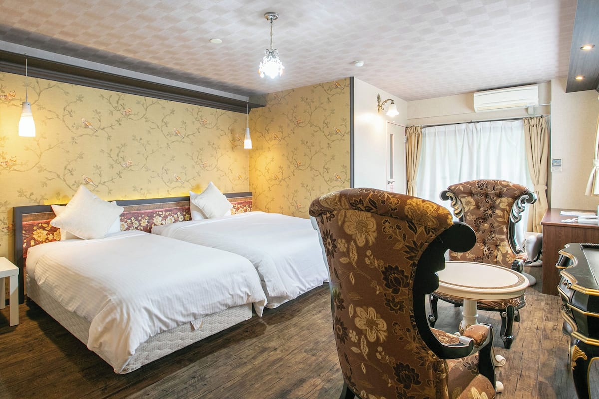 Quarto de hotel com duas camas, chão de madeira, poltronas com padrão floral, uma pequena mesa redonda e um lustre.