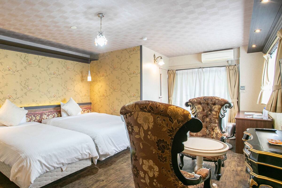 Quarto de hotel com duas camas, um espaço de estar com mesa e cadeiras, e um espelho grande.