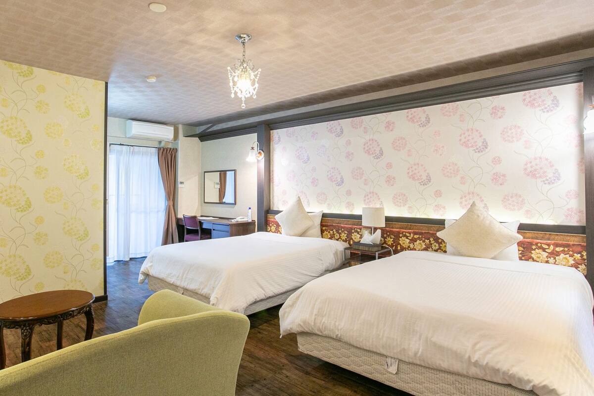 Quarto de hotel com duas camas, uma escrivaninha, uma cadeira, um candelabro e papel de parede floral.