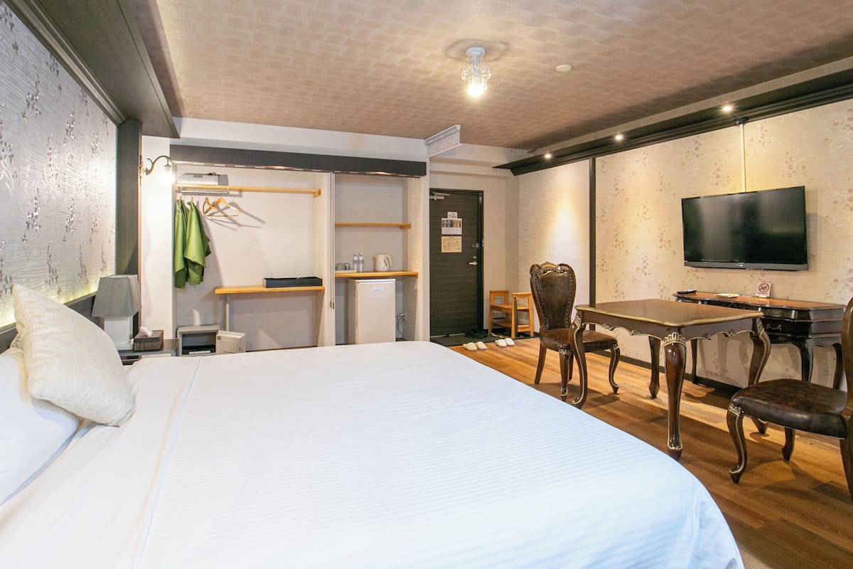 Quarto de hotel com uma cama grande, uma escrivaninha com cadeira, uma televisão e um guarda-roupa.