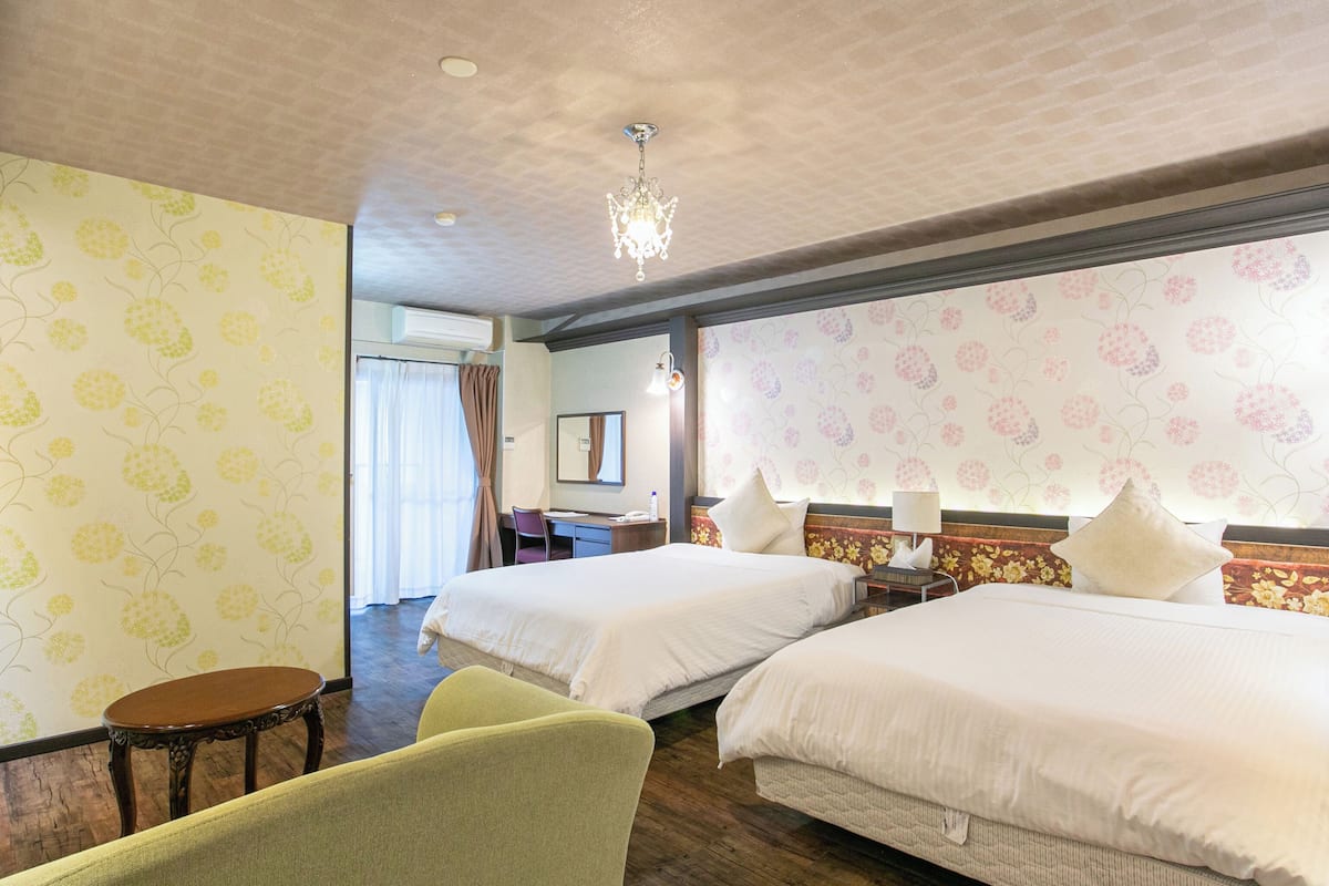 Quarto de hotel com duas camas, uma mesa, uma cadeira, um candelabro e papel de parede floral.