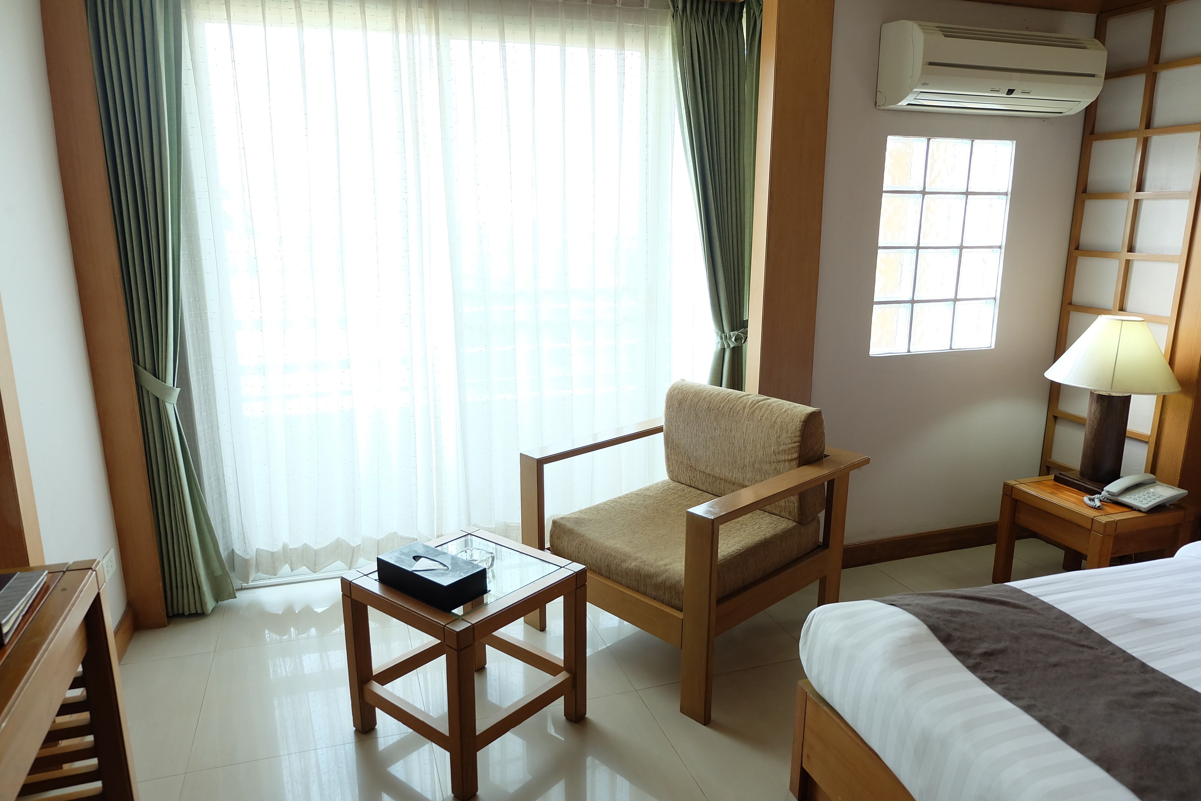 Quarto de hotel com uma cama, uma cadeira, uma mesinha com um livro, uma luminária e um aparelho de ar condicionado.